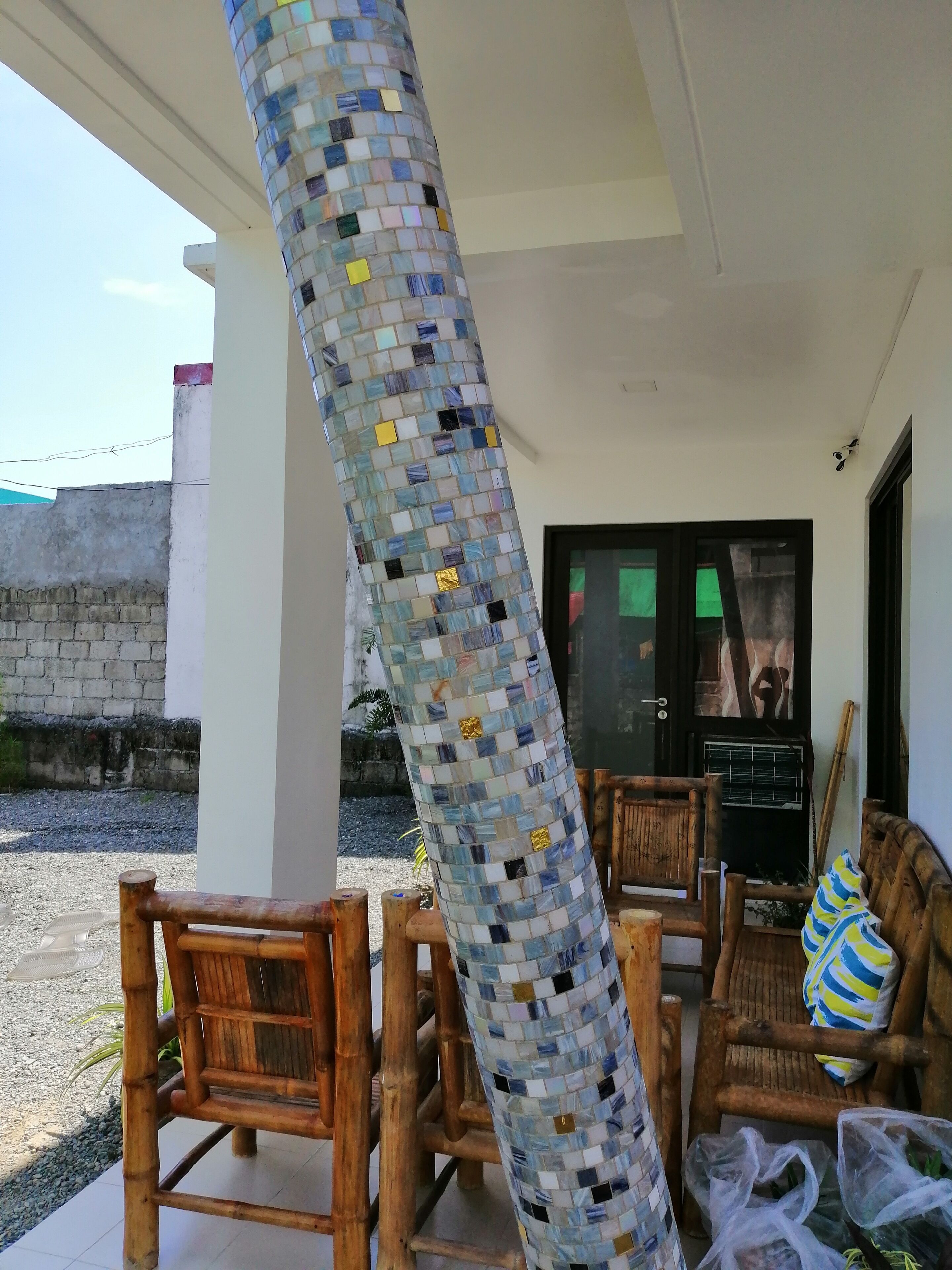 Uma área de estar ao ar livre coberta, com um pilar em mosaico, mobiliário de madeira e uma porta com cortina verde.