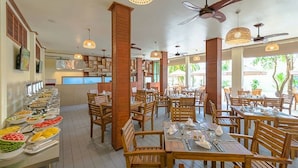 Restaurante com mesas e cadeiras de madeira, ventiladores de teto e um buffet com diversas opções de comida.