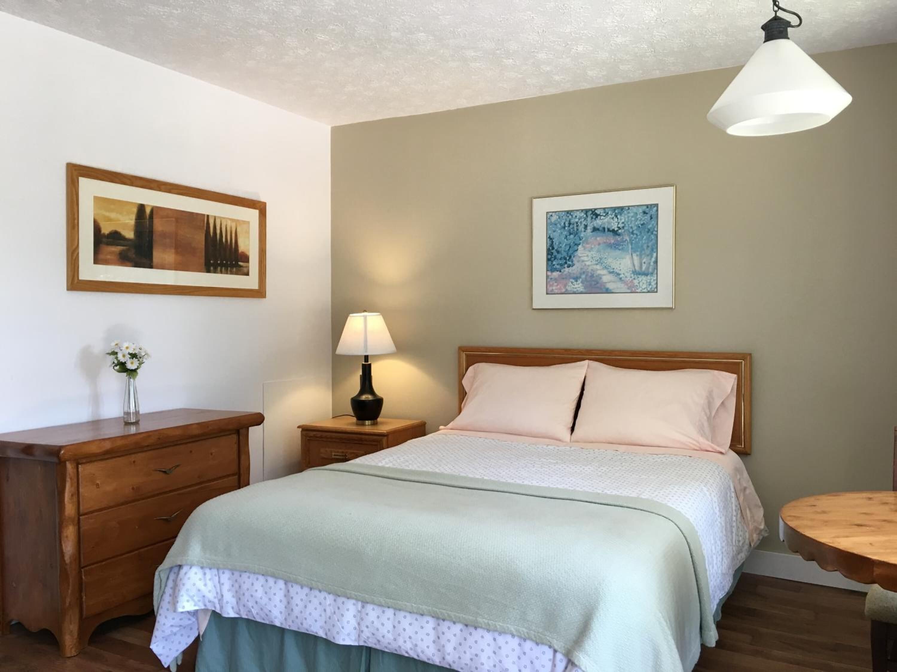 Quarto bem arrumado com cama, mesas de cabeceira, abajur e duas fotos emolduradas na parede.