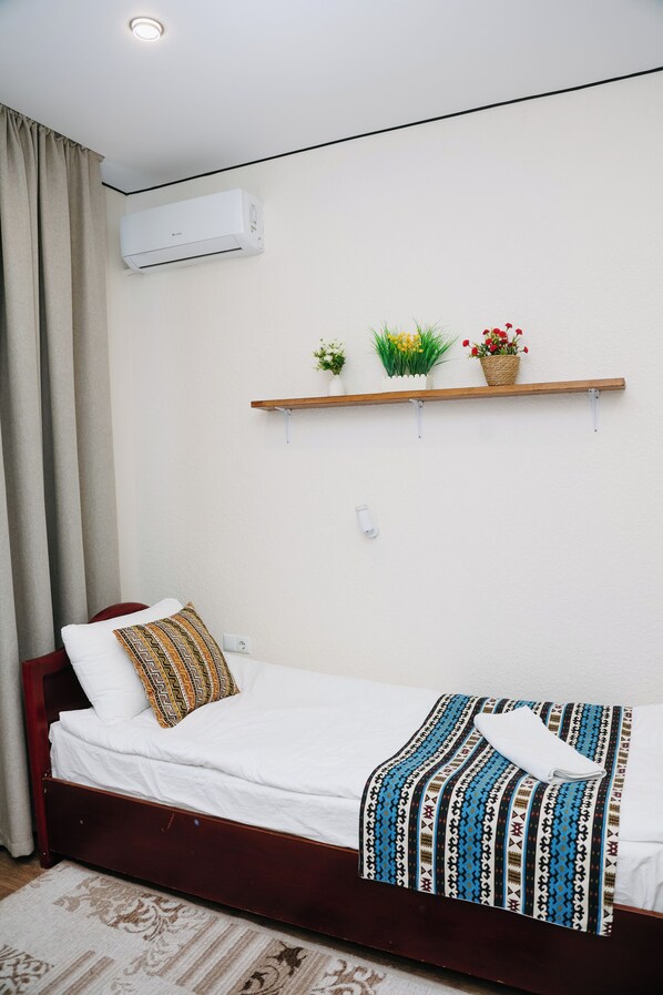 Quarto com cama, ar-condicionado na parede, prateleira com plantas e janela com cortinas.