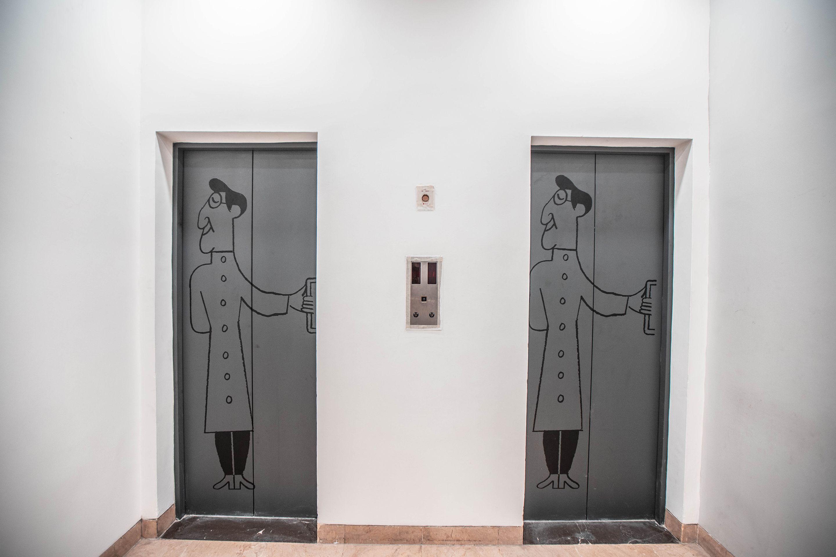 Duas portas de elevador com um desenho simples de uma pessoa segurando uma ferramenta, uma tomada e um interruptor de luz na parede entre elas.