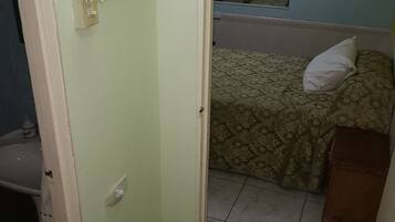Quarto com cama, mesa de cabeceira, luminária na parede e porta que dá para outro espaço.