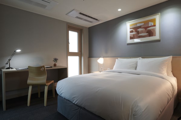 Quarto de hotel com uma cama grande, uma escrivaninha com cadeira, uma luminária de mesa e um quadro na parede.