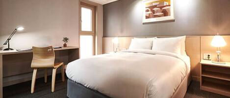 Quarto de hotel com uma cama grande, uma mesa com cadeira, uma mesinha de cabeceira com abajur e um quadro na parede.