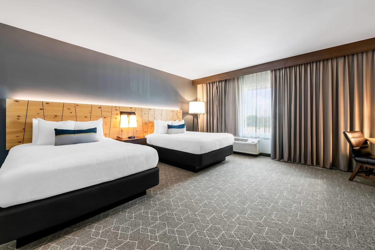 Quarto de hotel com duas camas, cabeceira de madeira, uma janela grande com cortinas e um tapete estampado.
