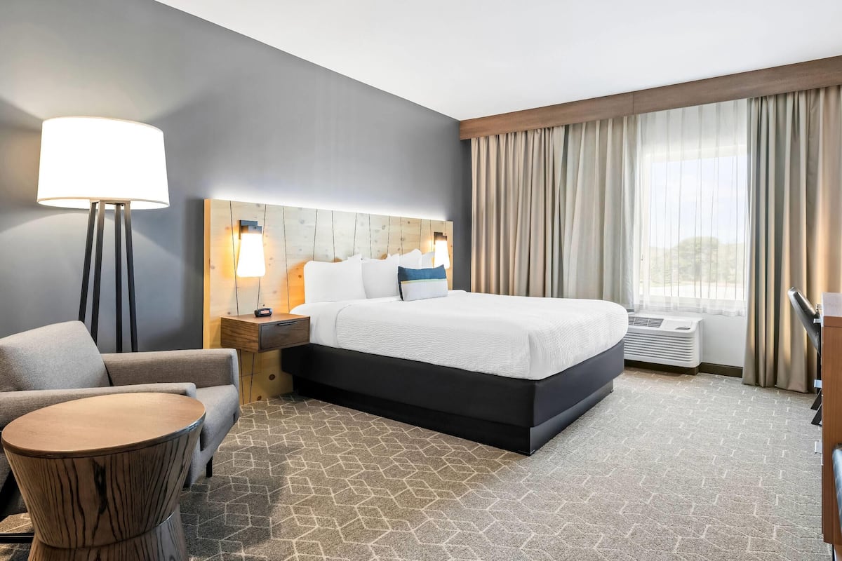 Quarto de hotel com uma cama, um sofá, uma mesa redonda, uma luminária, uma cabeceira e uma janela com cortinas.