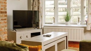 Sala de estar com televisão, mesa de centro branca, sofá e janela com cortinas.