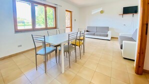 Uma sala de estar ampla com uma mesa de jantar e cadeiras, um sofá e uma televisão fixada na parede.