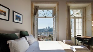 Quarto de hotel com cama, escrivaninha e vista para a cidade pelas janelas abertas.