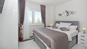 Quarto moderno com cama grande, mesas de cabeceira, luminária na parede e vista para o exterior pela janela.