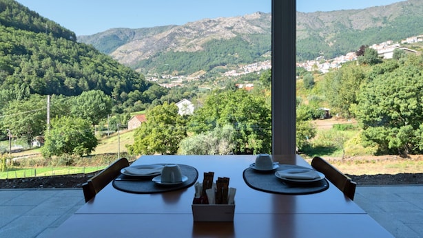 Uma mesa de jantar com pratos e talheres, com uma paisagem montanhosa e uma aldeia ao fundo.