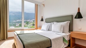 Quarto de hotel com uma cama grande, uma mesa de cabeceira de madeira e vista para uma cidade através de uma janela ampla.