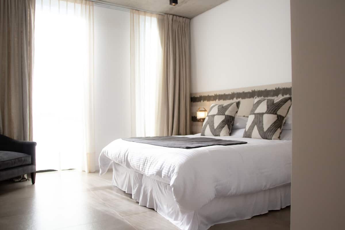 Cama bem arrumada com edredom branco e manta cinza, uma poltrona cinza e uma janela grande com cortinas leves.