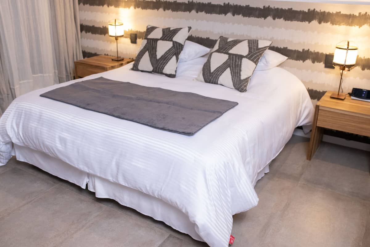 Cama bem arrumada com um cobertor cinza, duas luminárias de cabeceira e papel de parede estampado.