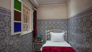 Quarto com cama, paredes estampadas, janela com grades decorativas de ferro e uma pequena mesa com uma planta.