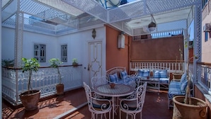 Pátio iluminado pelo sol com mesa e cadeiras de metal branco, um banco acolchoado e plantas em vasos.