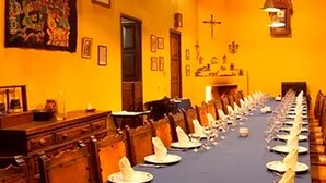 Uma longa mesa de jantar posta para uma refeição, com cadeiras em ambos os lados, uma cruz na parede e um quadro sobre a mesa.