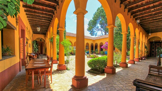 Um pátio aberto com uma mesa e cadeiras de madeira, cercado por colunas e arcos amarelos, e uma placa com a inscrição 'RESTAURANTE'.
