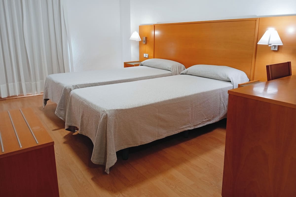Quarto de hotel com duas camas de solteiro, cabeceiras de madeira, abajures, uma escrivaninha de madeira e piso de madeira.