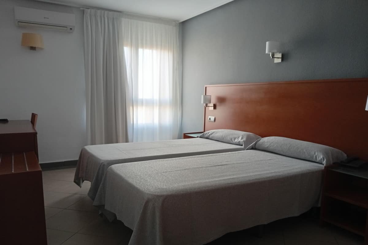 Quarto de hotel com duas camas, cabeceira de madeira, mesa de cabeceira e escrivaninha com abajur.