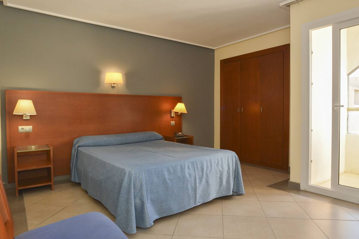 Quarto de hotel com uma cama de casal, duas luminárias de cabeceira, um painel de cama de madeira, uma mesa de cabeceira e um guarda-roupa.