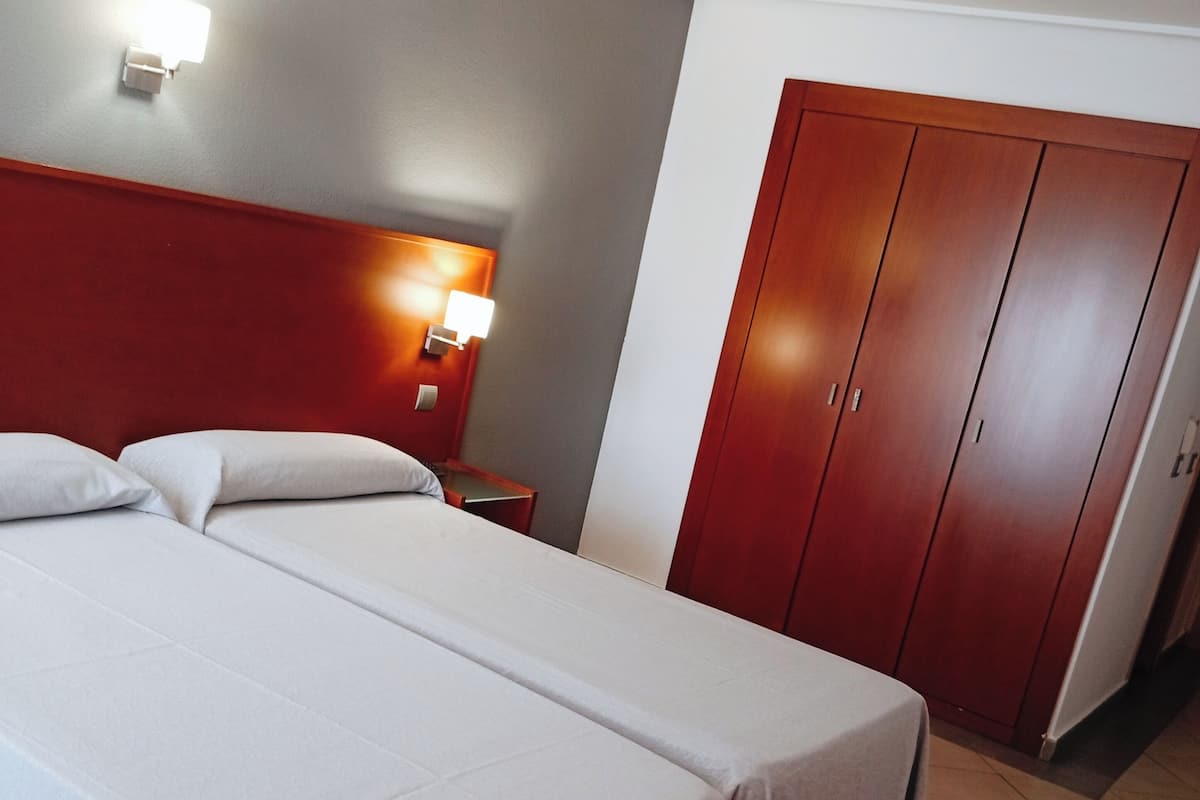 Quarto de hotel com uma cama grande, uma mesa de cabeceira com abajur, um guarda-roupa e um tapete vermelho.