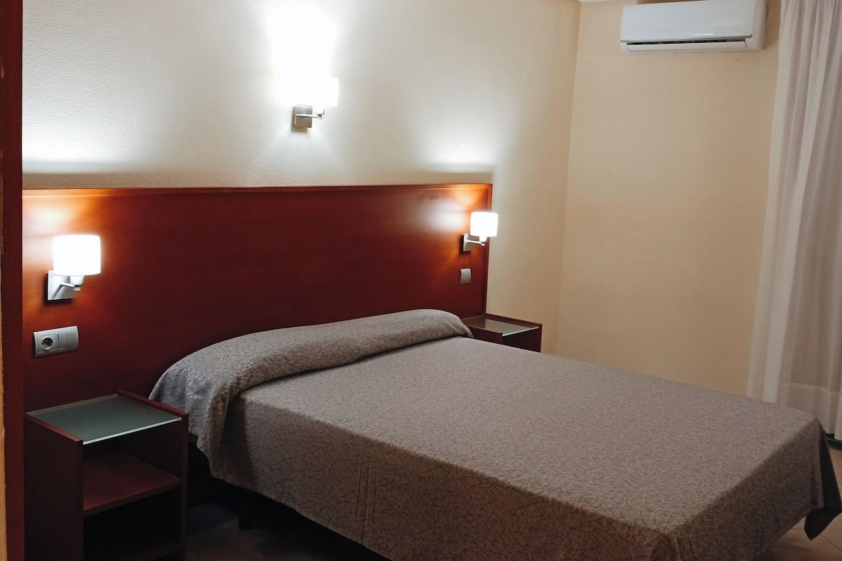 Quarto de hotel com uma cama de solteiro, duas mesas de cabeceira, um ar condicionado fixado na parede e duas luzes também fixadas na parede.