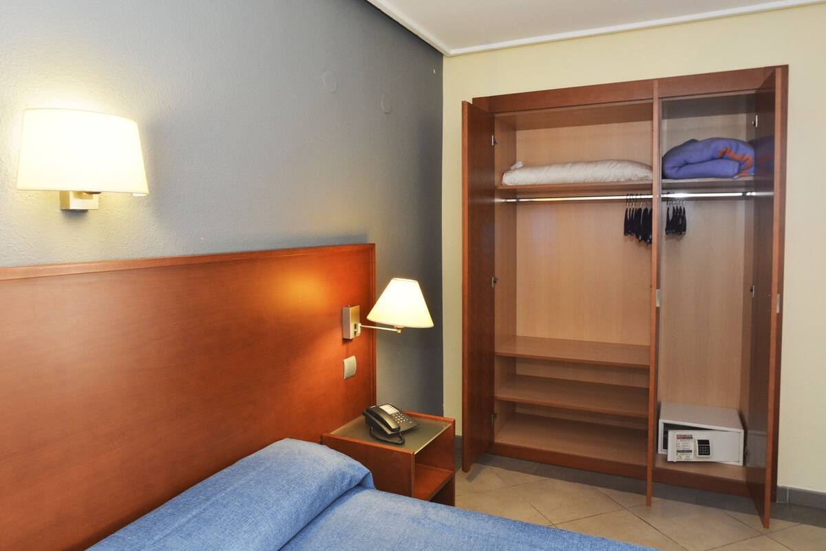 Quarto de hotel com cama, luminária de cabeceira, painel de madeira e um closet espaçoso com roupas penduradas.