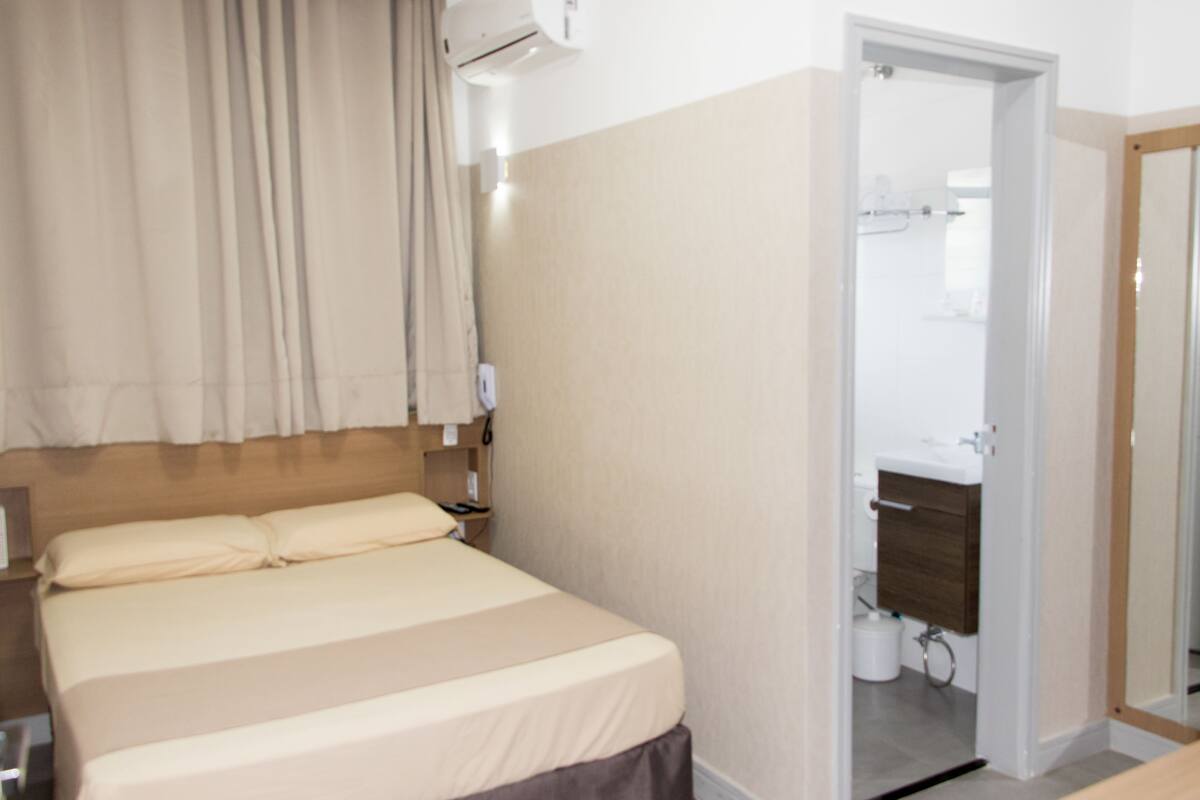 Quarto de hotel com cama, mesa de cabeceira, unidade de ar condicionado e banheiro visível por uma porta aberta.