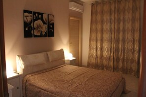 Quarto com cama, mesa de cabeceira, ar condicionado fixado na parede e cortina.