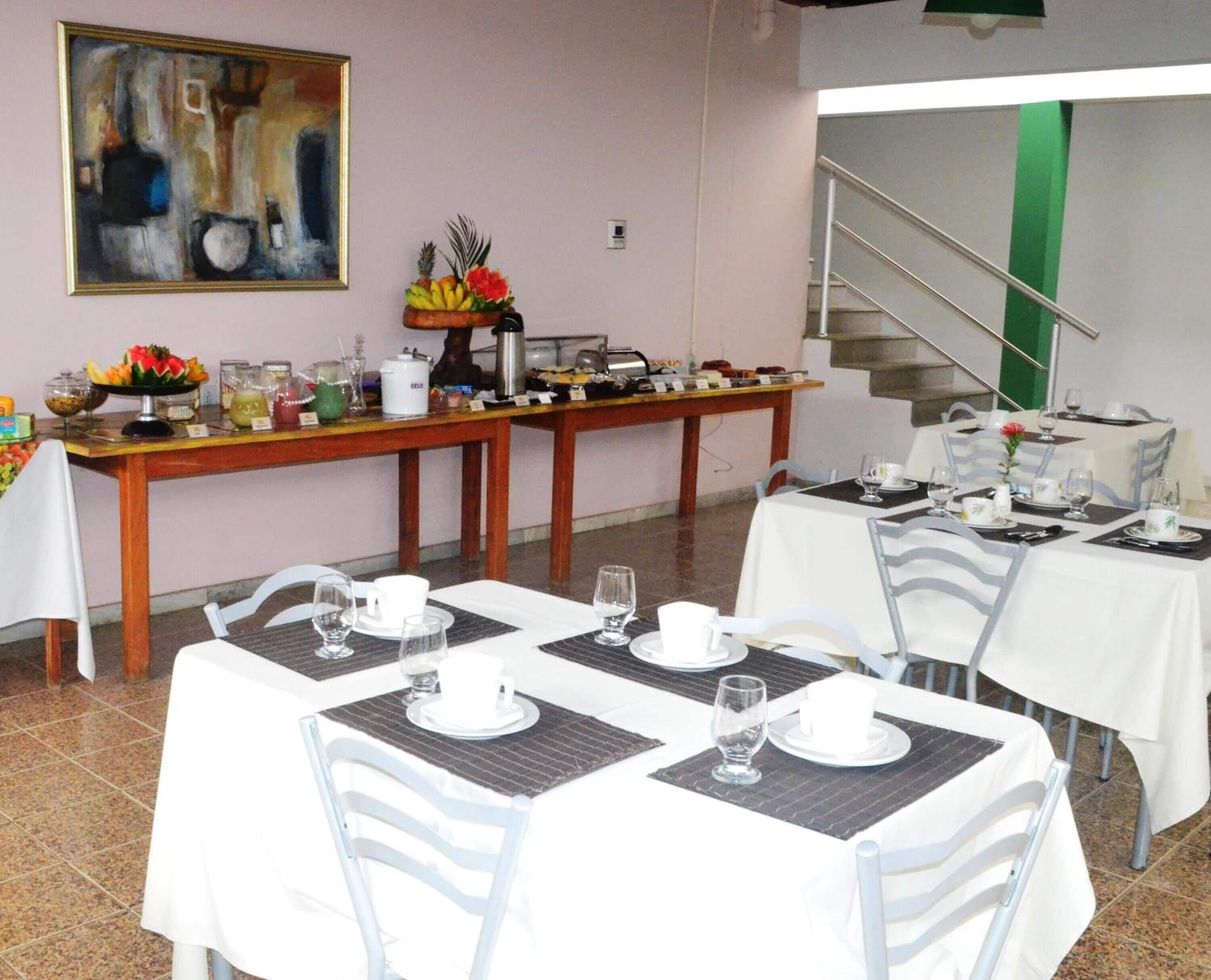 Uma área de refeições com mesas preparadas para o café da manhã, uma mesa com frutas e uma escada ao fundo.