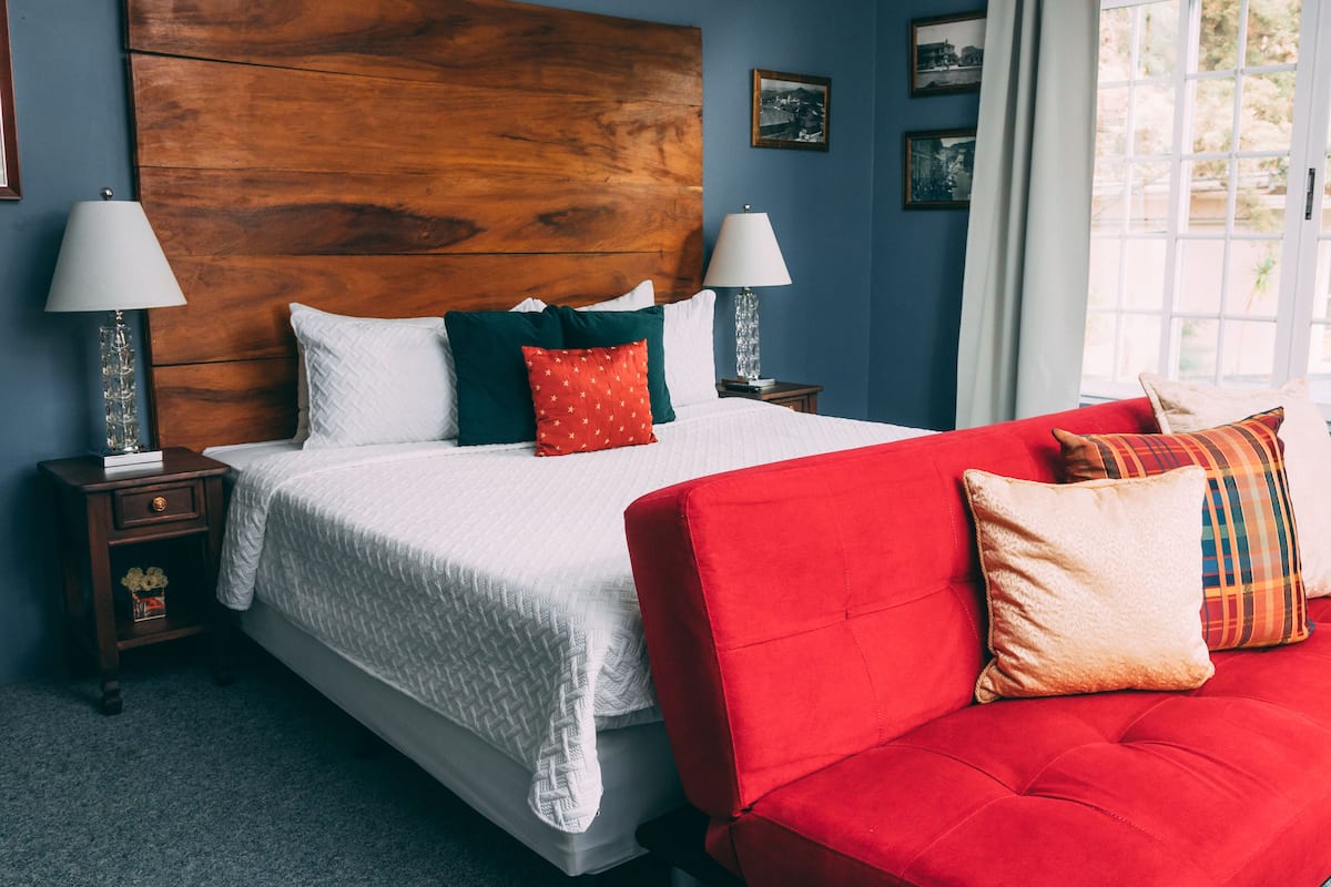 Quarto com cabeceira de madeira, cama com roupa de cama branca, duas mesas de cabeceira com luminárias, um sofá vermelho e uma janela com cortinas.