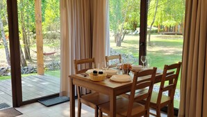 Uma área de refeições com uma mesa posta para quatro pessoas, janelas amplas com cortinas e vista para um jardim.