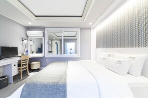 Quarto de hotel moderno com uma cama grande, uma escrivaninha, uma cadeira, um banquinho pequeno e um espelho.