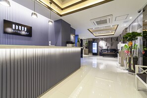 Uma área de recepção de hotel moderna, com balcão de recepção, espaço de estar com sofá e mesa de centro, e uma grande planta em vaso.