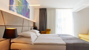 Quarto de hotel com uma cama grande, uma mesinha de cabeceira com abajur, uma cadeira e um quadro na parede.