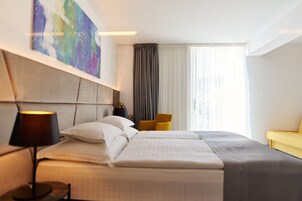 Quarto de hotel com uma cama grande, uma mesinha de cabeceira com abajur, uma cadeira e um quadro na parede.