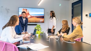 Sala de conferências com uma tela grande exibindo uma paisagem desértica, um quadro branco com gráficos e texto, e um relógio marcando 15:35.