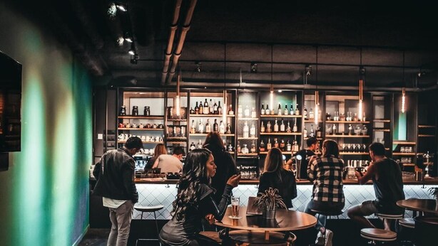 Um bar com mesas e cadeiras redondas, prateleiras com garrafas e uma parede verde decorativa.