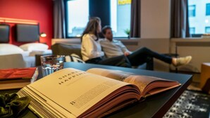Quarto de hotel com uma janela grande, um sofá e uma mesa com um livro aberto.