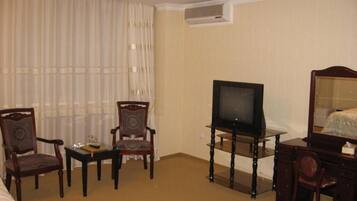 Quarto de hotel com cama, televisão sobre suporte, escrivaninha com cadeira e duas poltronas.