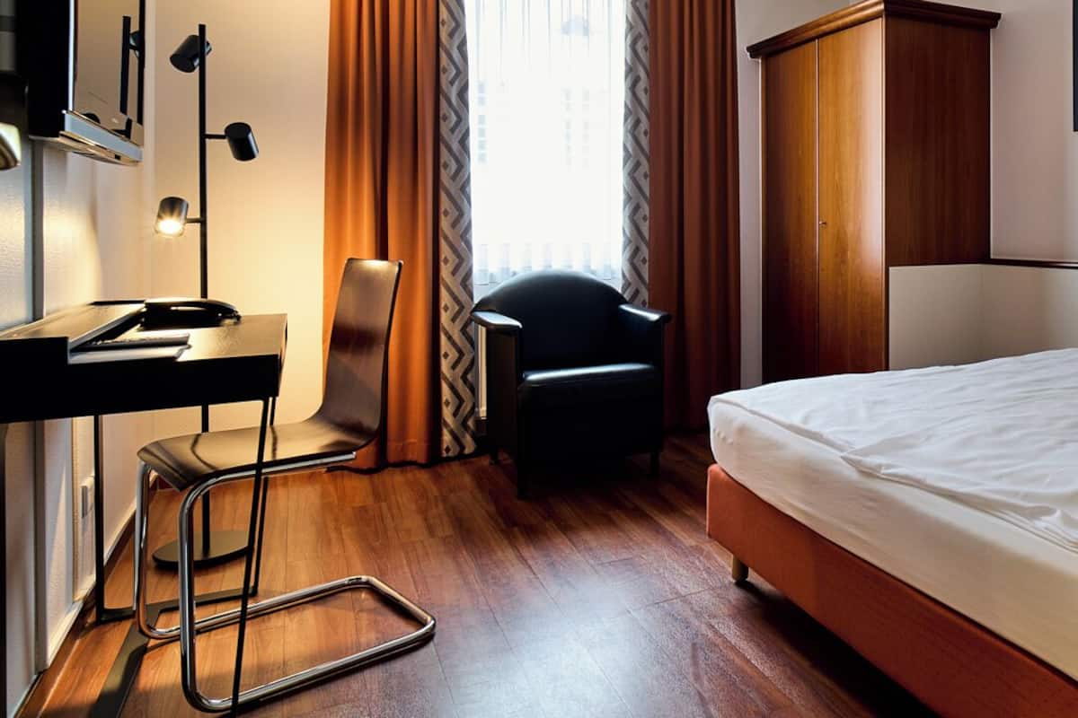 Quarto de hotel com cama, escrivaninha, cadeira, guarda-roupa e uma janela grande com cortinas.