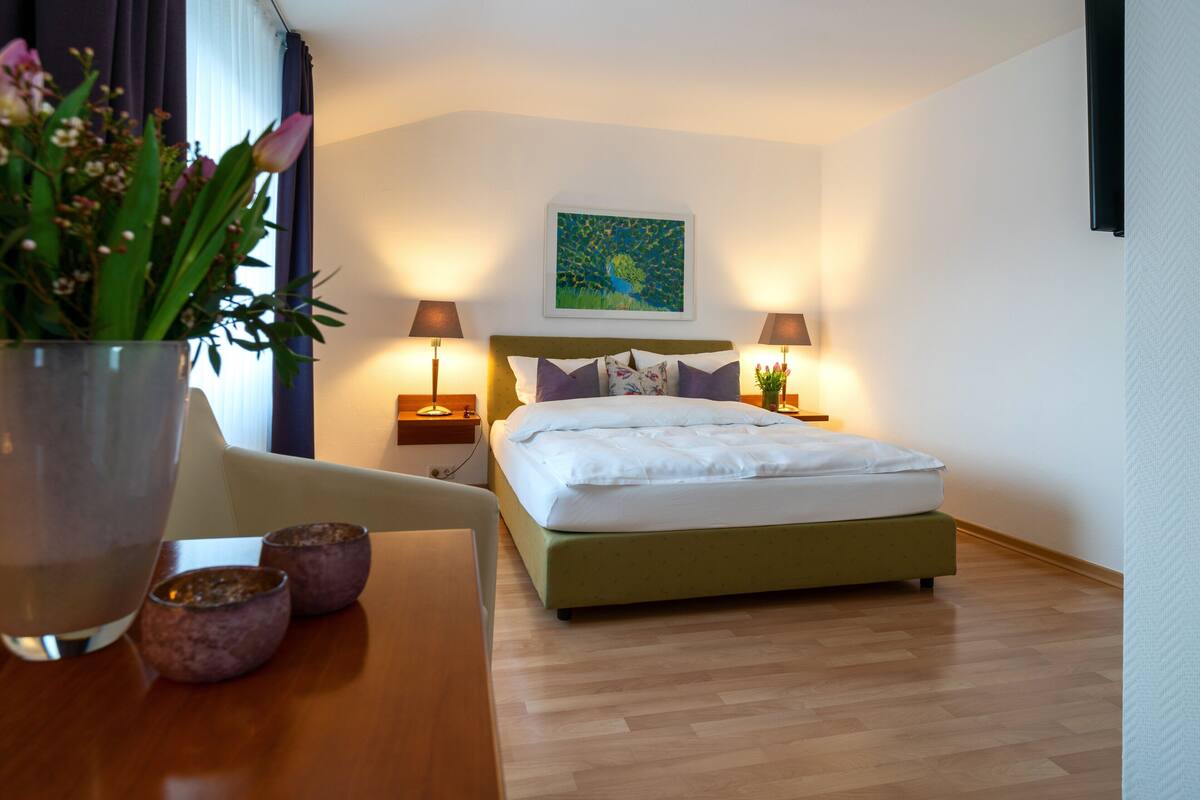 Quarto de hotel com cama, abajures, um quadro na parede e um vaso com flores na mesa.