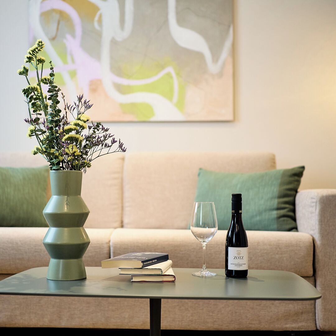 Uma sala de estar moderna com um vaso de flores verdes sobre uma mesa de vidro, uma garrafa de vinho e um copo de vinho.