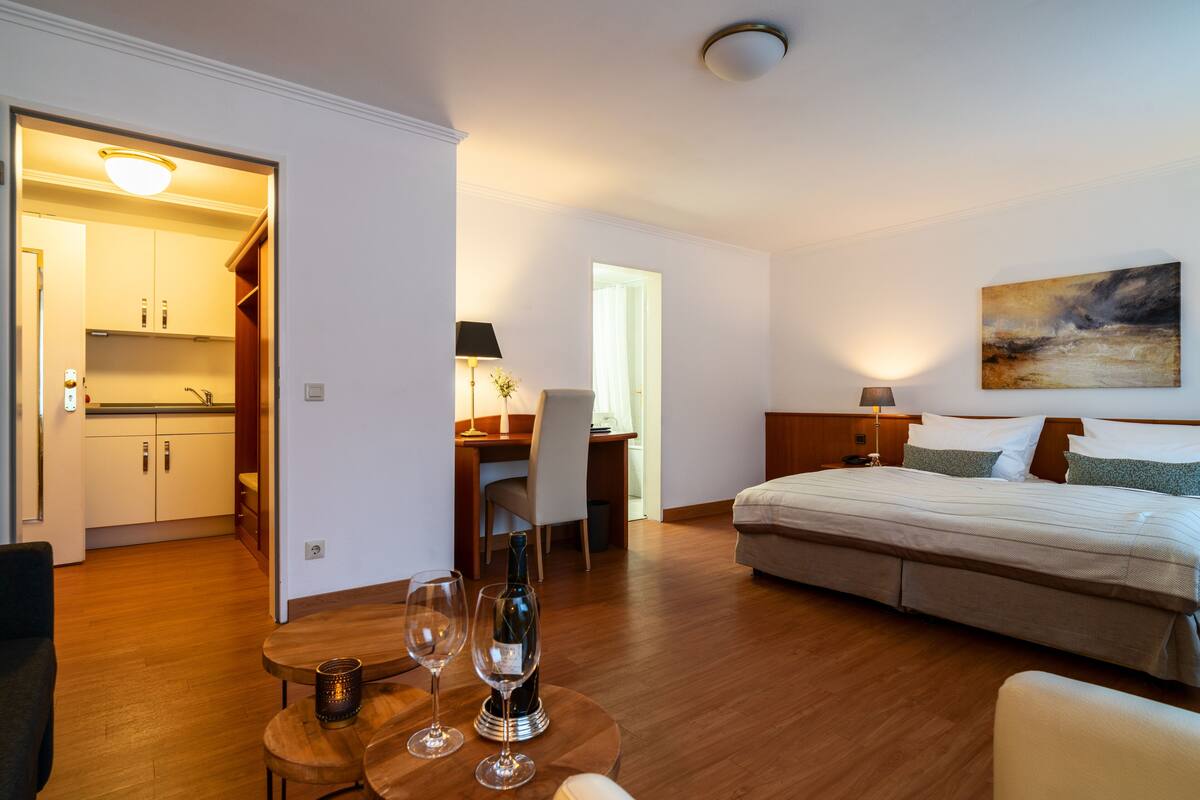 Quarto de hotel com cama, escrivaninha, cadeira, mesa com copos de vinho e um quadro na parede.