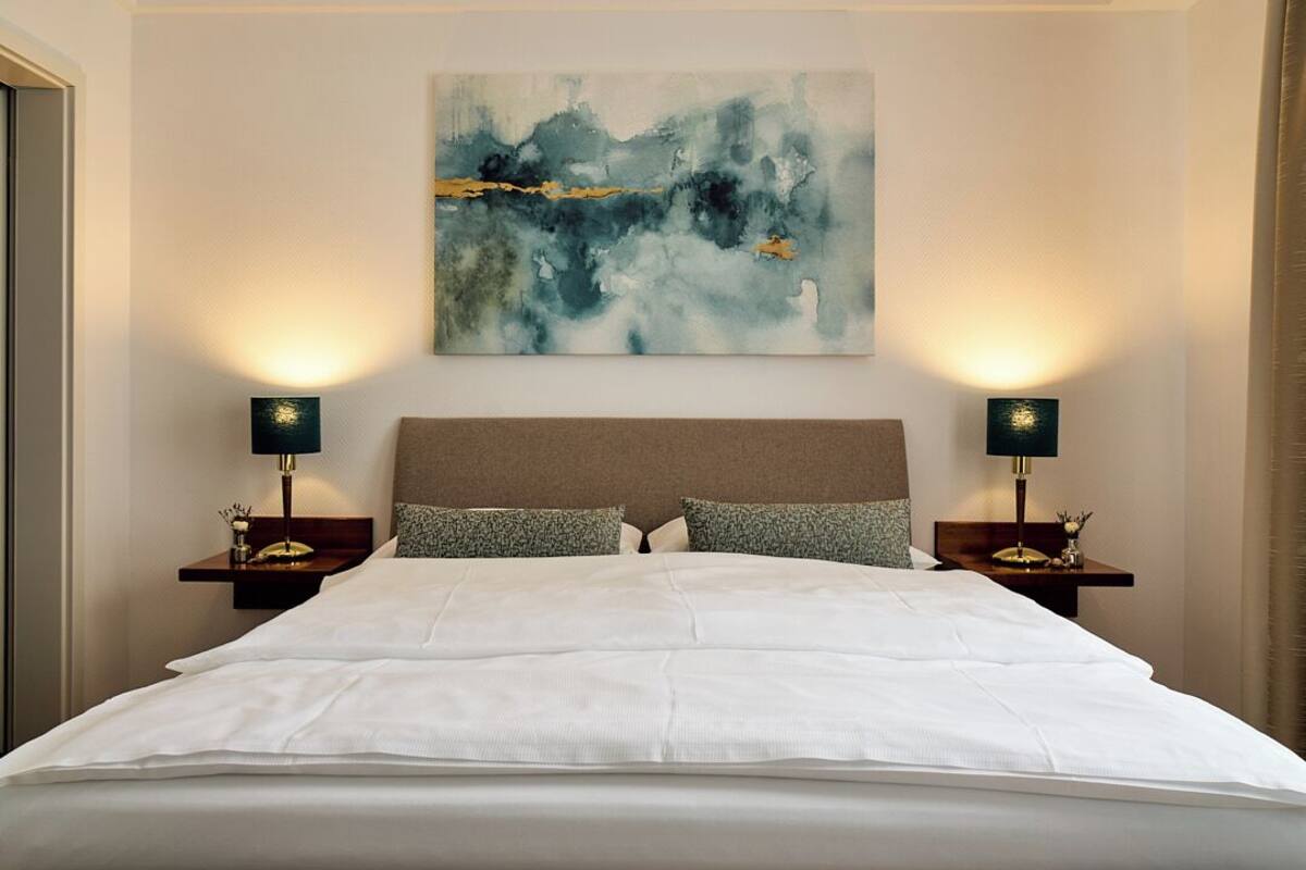 Quarto de hotel com uma cama grande, duas mesas de cabeceira com luminárias, um quadro abstrato na parede e vista para a cidade.