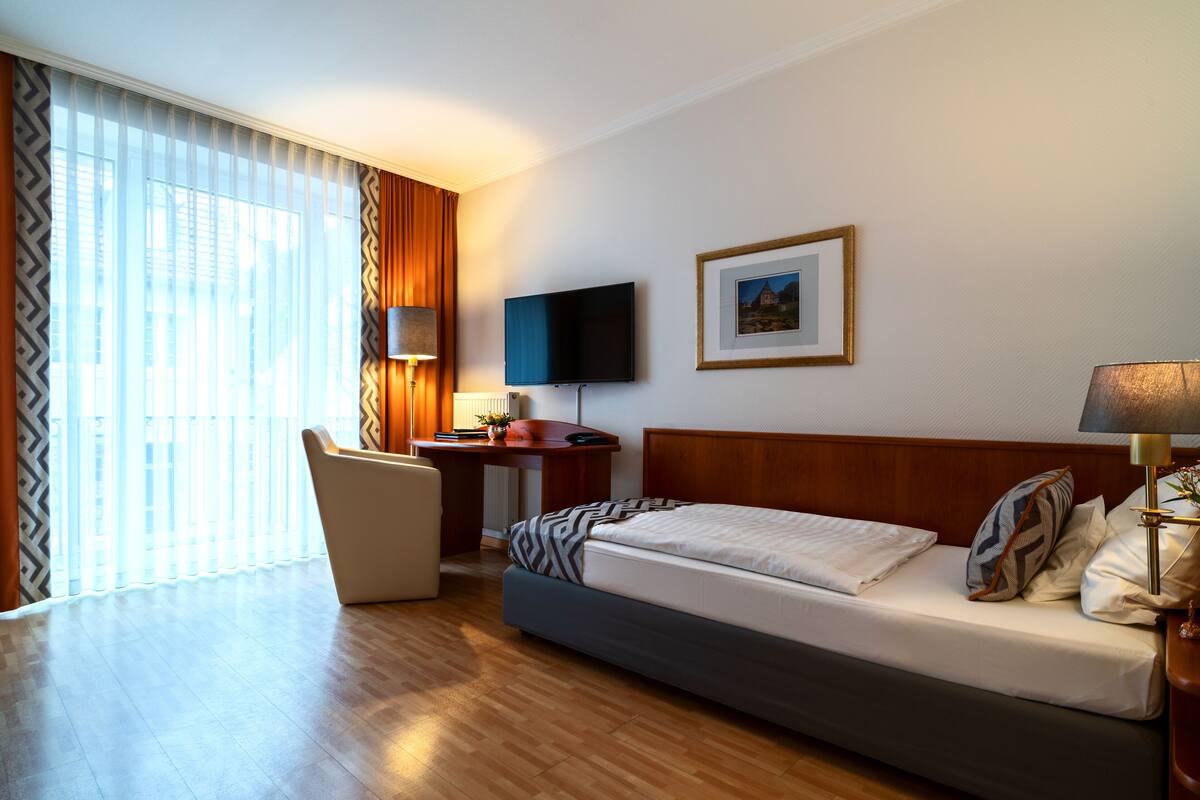 Quarto de hotel com cama, escrivaninha, cadeira, televisão e janela com cortinas.
