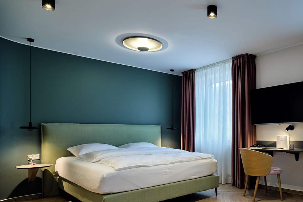 Quarto de hotel moderno com uma cama grande, uma escrivaninha com cadeira, uma televisão e uma luminária no teto.