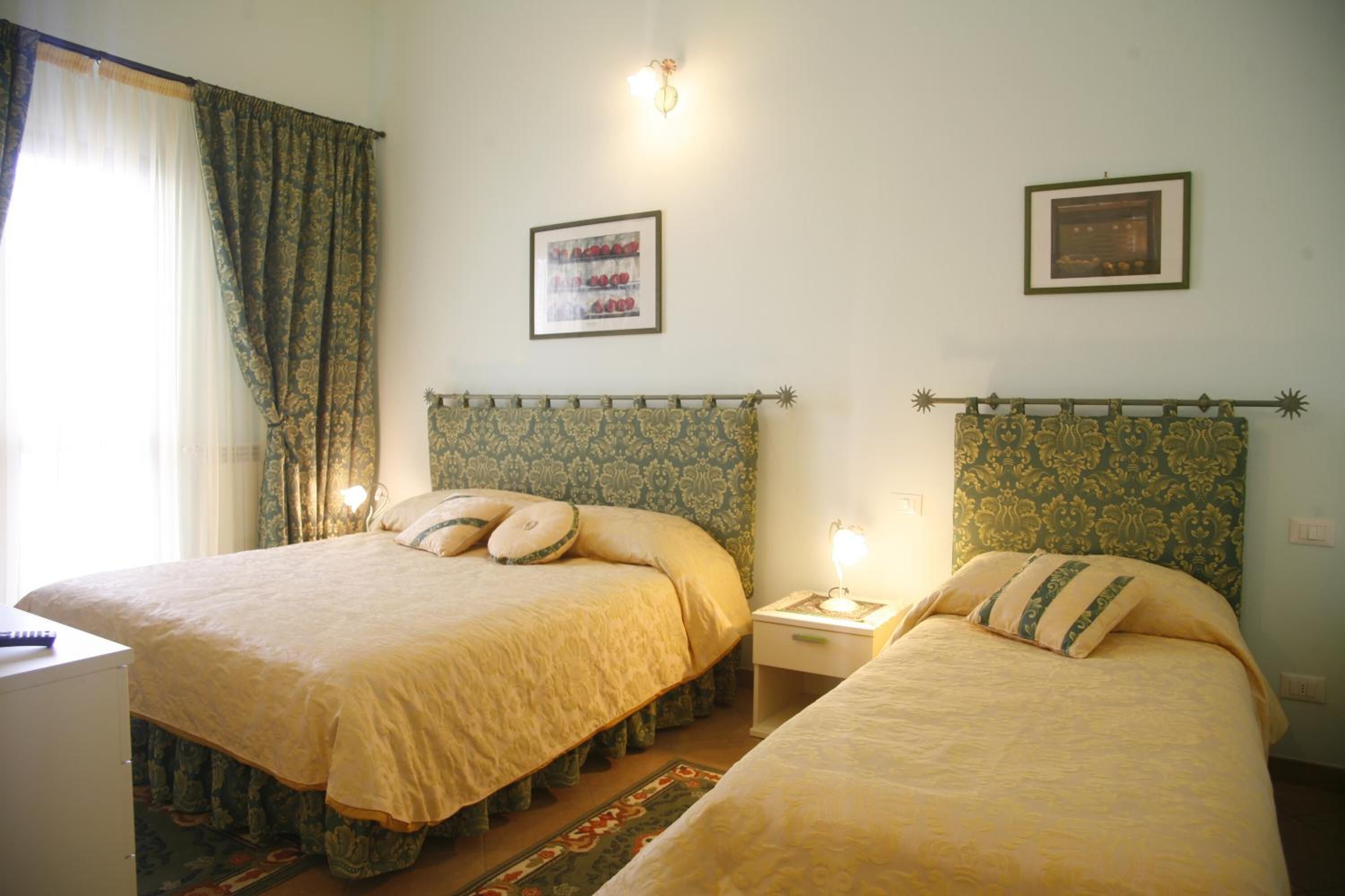 Quarto de hotel com duas camas, uma mesinha de cabeceira com abajur, janela com cortinas e duas fotos emolduradas na parede.