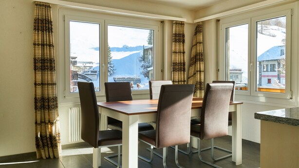 Uma sala de jantar com mesa e cadeiras, amplas janelas com vista para uma montanha nevada e edifícios.